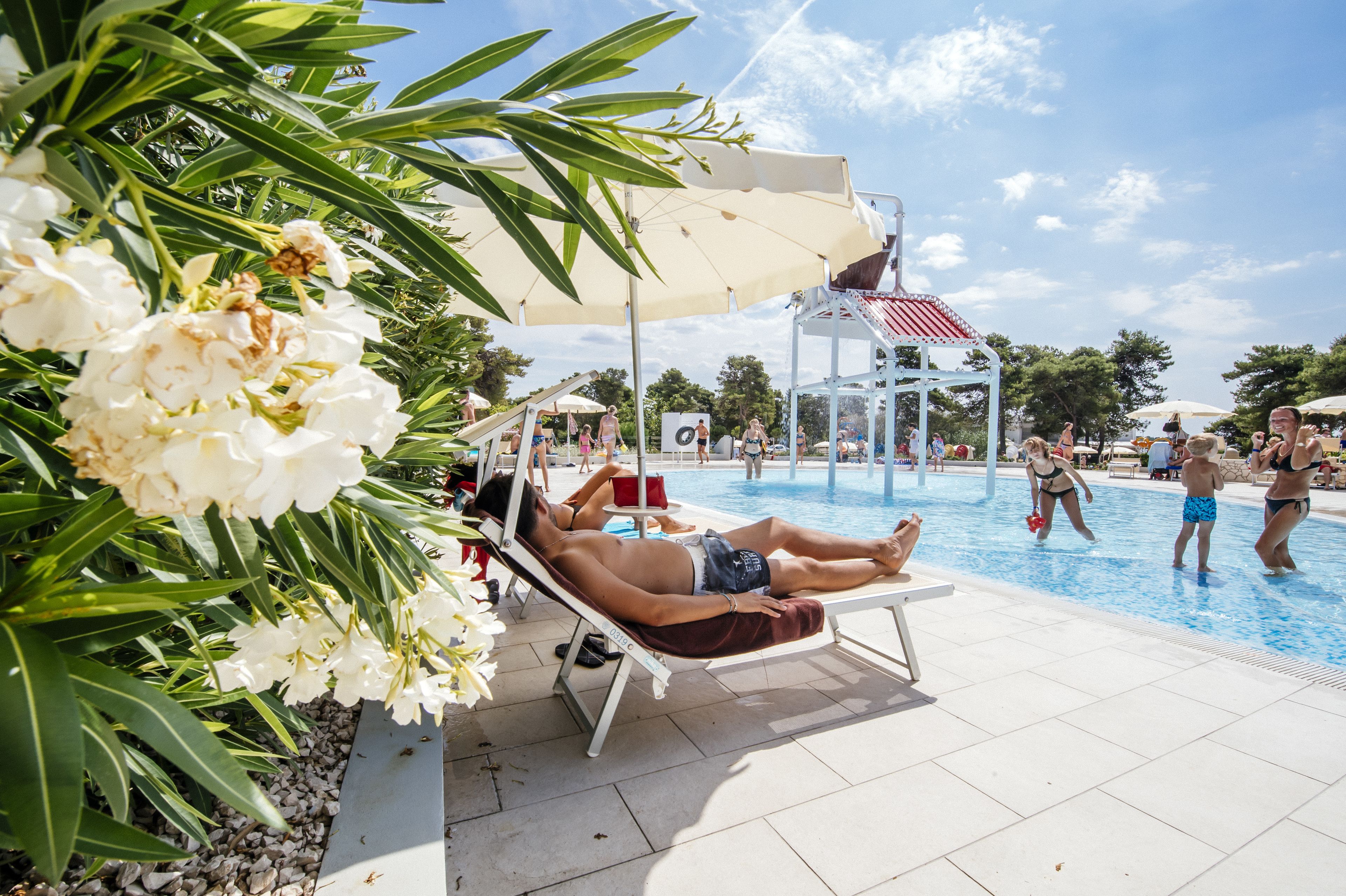 Uma área à beira da piscina com espreguiçadeiras e guarda-sóis, um tobogã e um pavilhão.