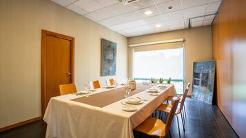 Uma sala de conferências com uma mesa comprida preparada para uma reunião, equipada com cadeiras, uma tela de projetor e uma grande janela que proporciona iluminação natural.