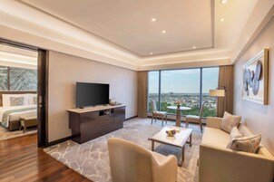 Quarto de hotel moderno com uma cama grande, televisão, área de refeições e vista para a cidade.