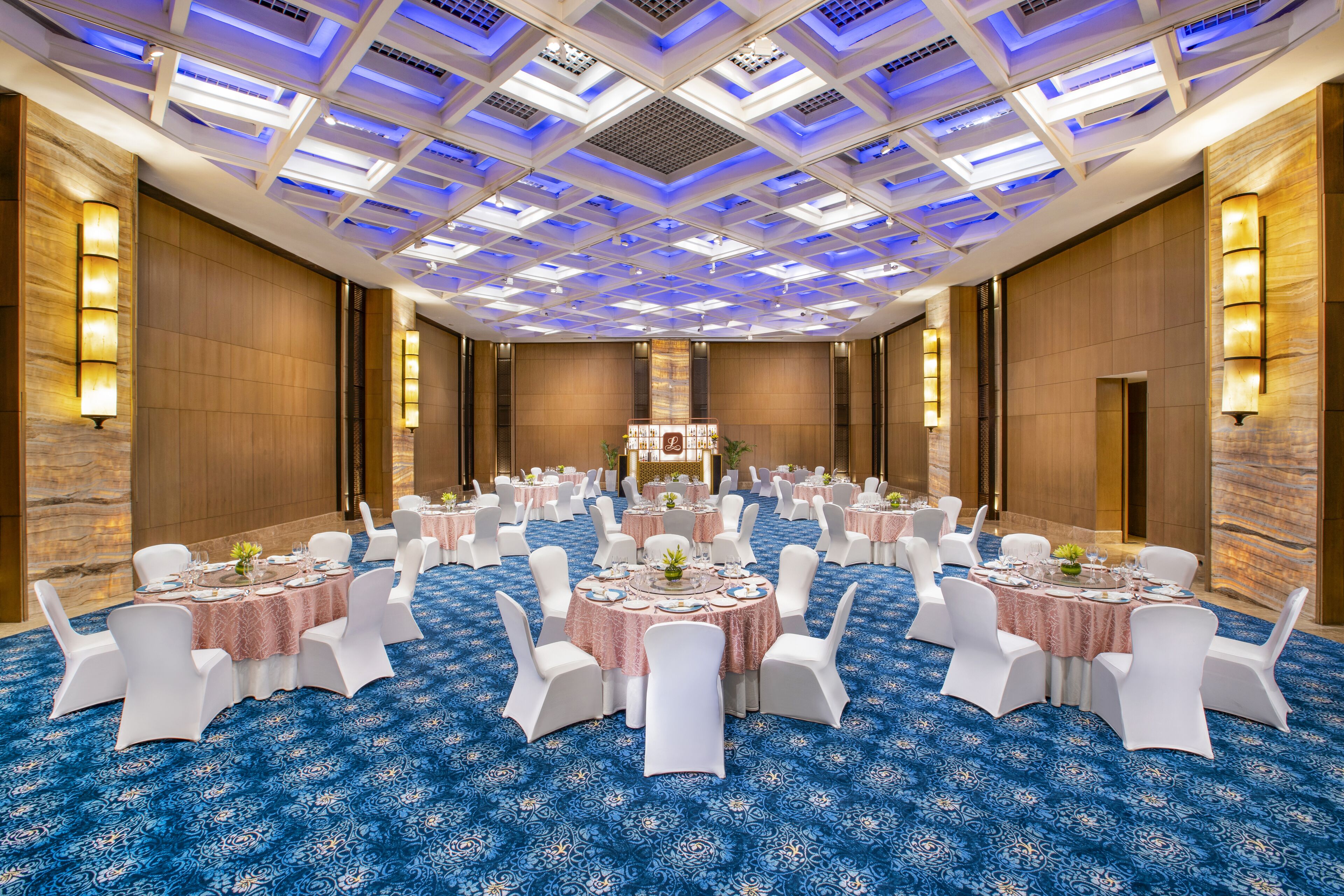 Uma sala de banquetes com mesas redondas, cadeiras brancas e um tapete azul estampado.