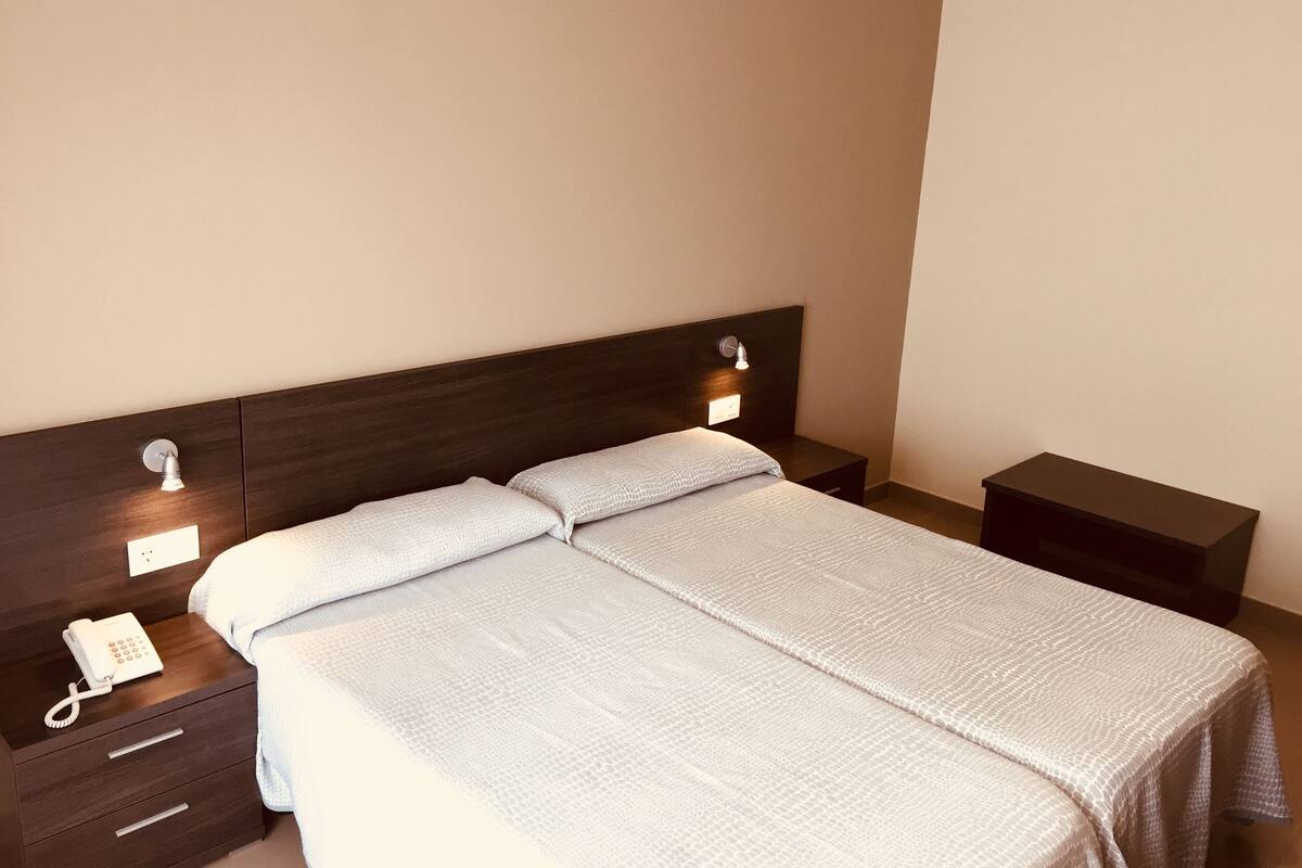 Uma cama bem arrumada com cabeceira de madeira, mesa de cabeceira com telefone e um criado-mudo.
