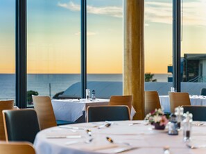 Restaurante com mesas preparadas para refeições, grandes janelas e vista para o mar.
