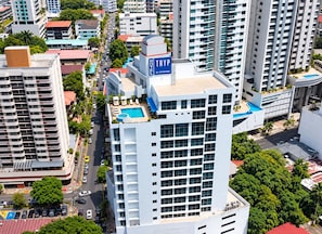 Um hotel com muitos andares e piscina no terraço, rodeado por edifícios urbanos.