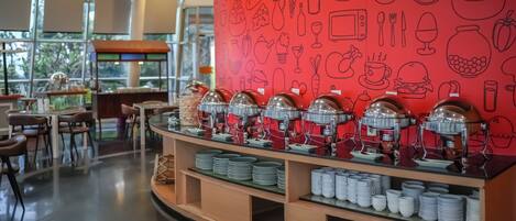 Uma área de buffet com vários distribuidores de comida, um balcão com pratos empilhados e uma parede vermelha com ilustrações de alimentos.