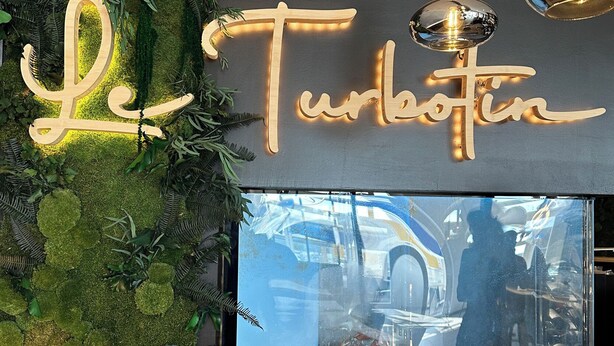 Interior de restaurante com um letreiro indicando "Le Turkoftin", uma parede de madeira com plantas e um aquário grande com peixes e um caranguejo.