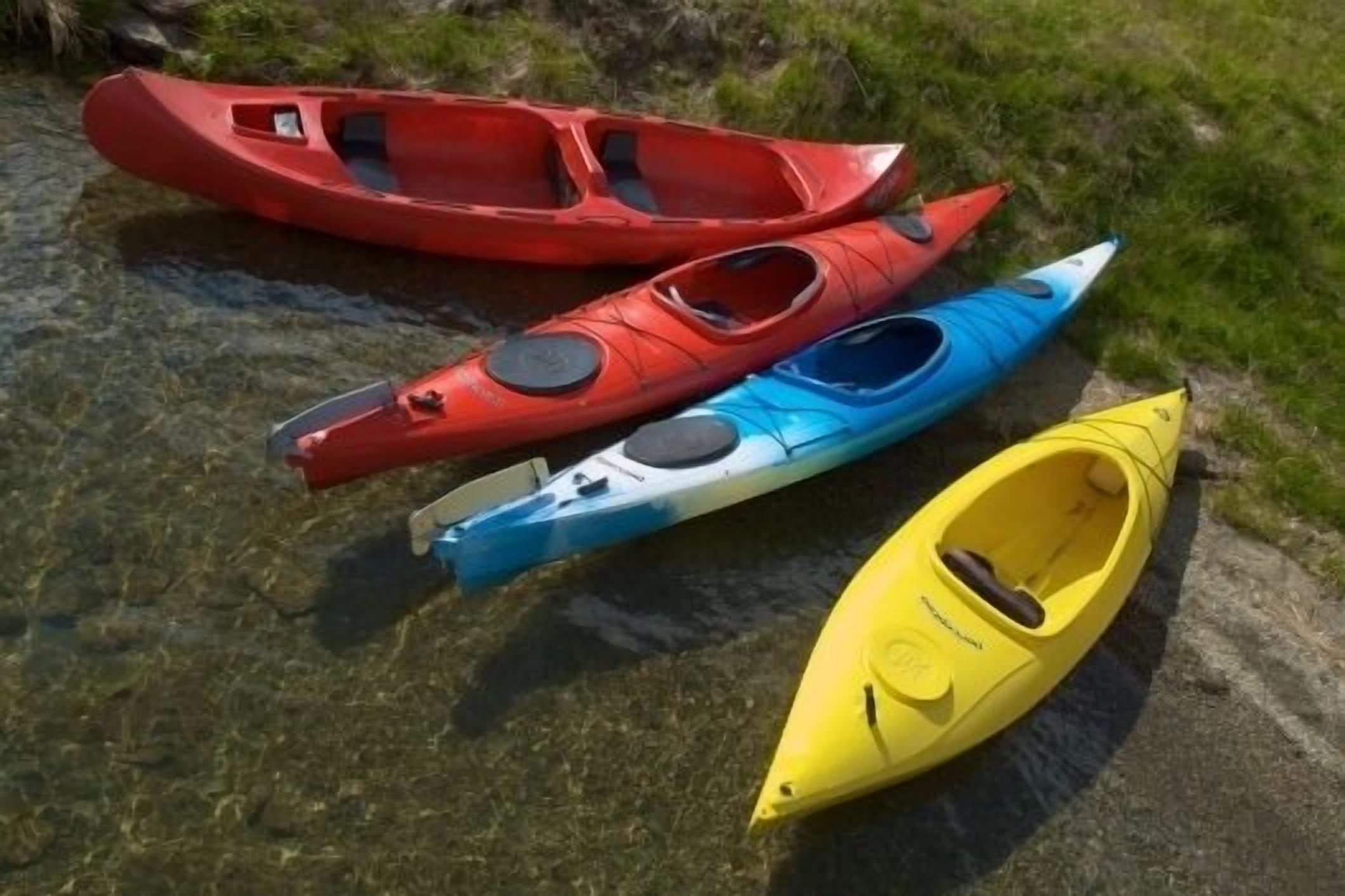 Três caiaques de cores diferentes (vermelho, azul e amarelo) colocados numa área gramada perto de um corpo d'água.