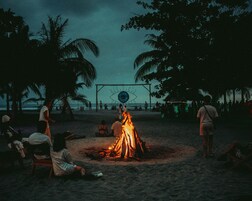 Cena de praia com uma fogueira no centro, cercada por pessoas. Palmeiras e um guarda-sol de praia são visíveis ao fundo.