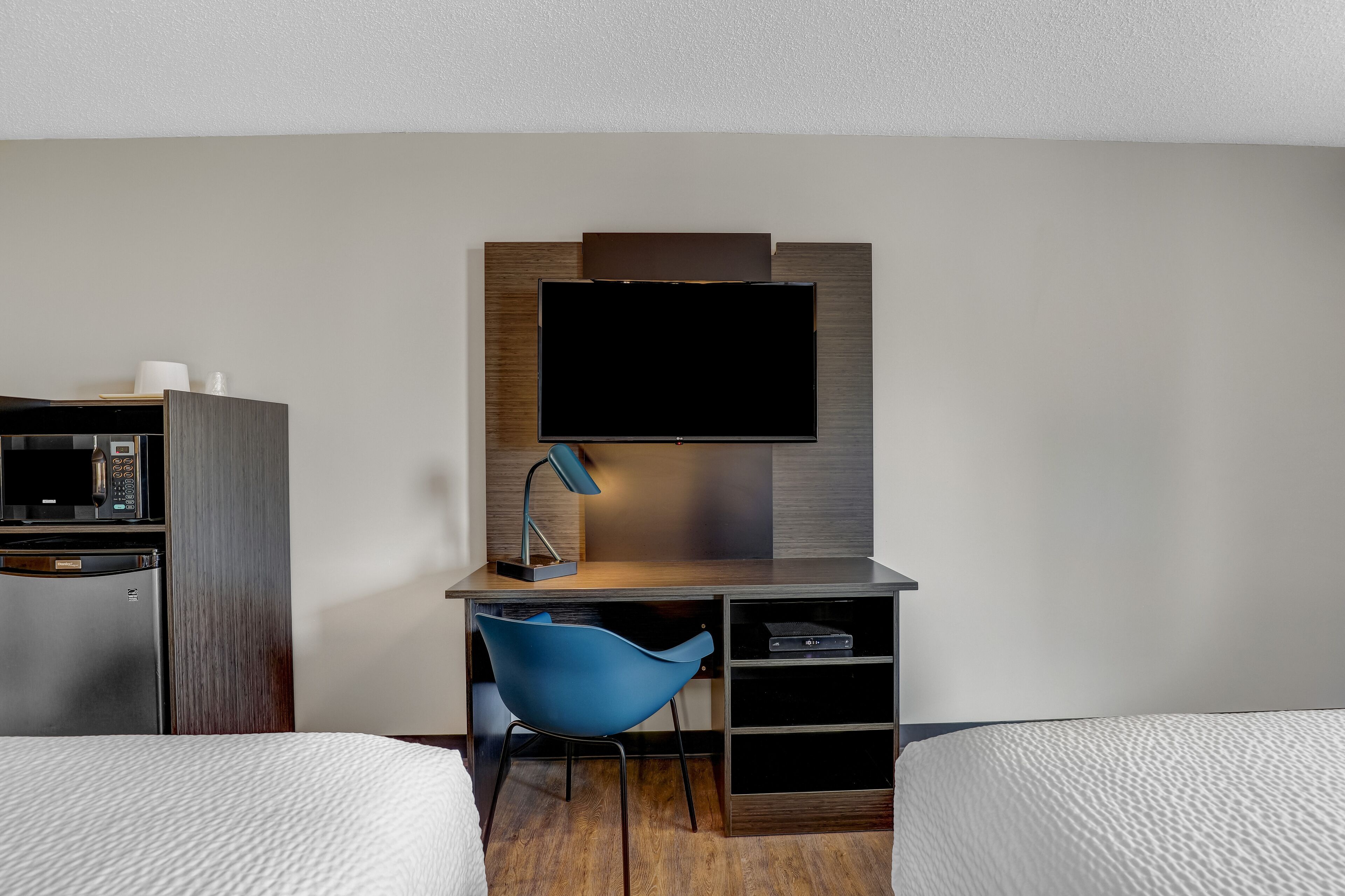 Quarto de hotel moderno com uma escrivaninha de madeira, uma cadeira azul, uma televisão de tela plana, um micro-ondas e um frigorífico.