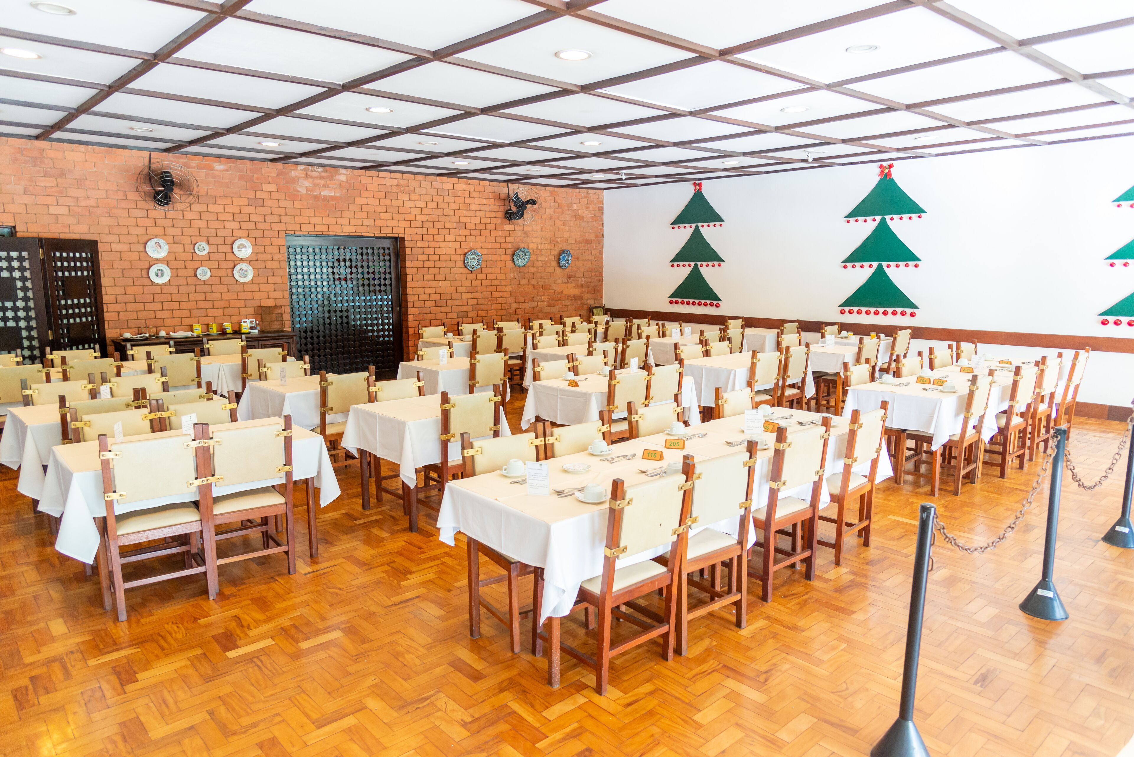 Um amplo salão de festas com várias mesas e cadeiras, uma parede de tijolos e árvores de Natal decorativas na parede.