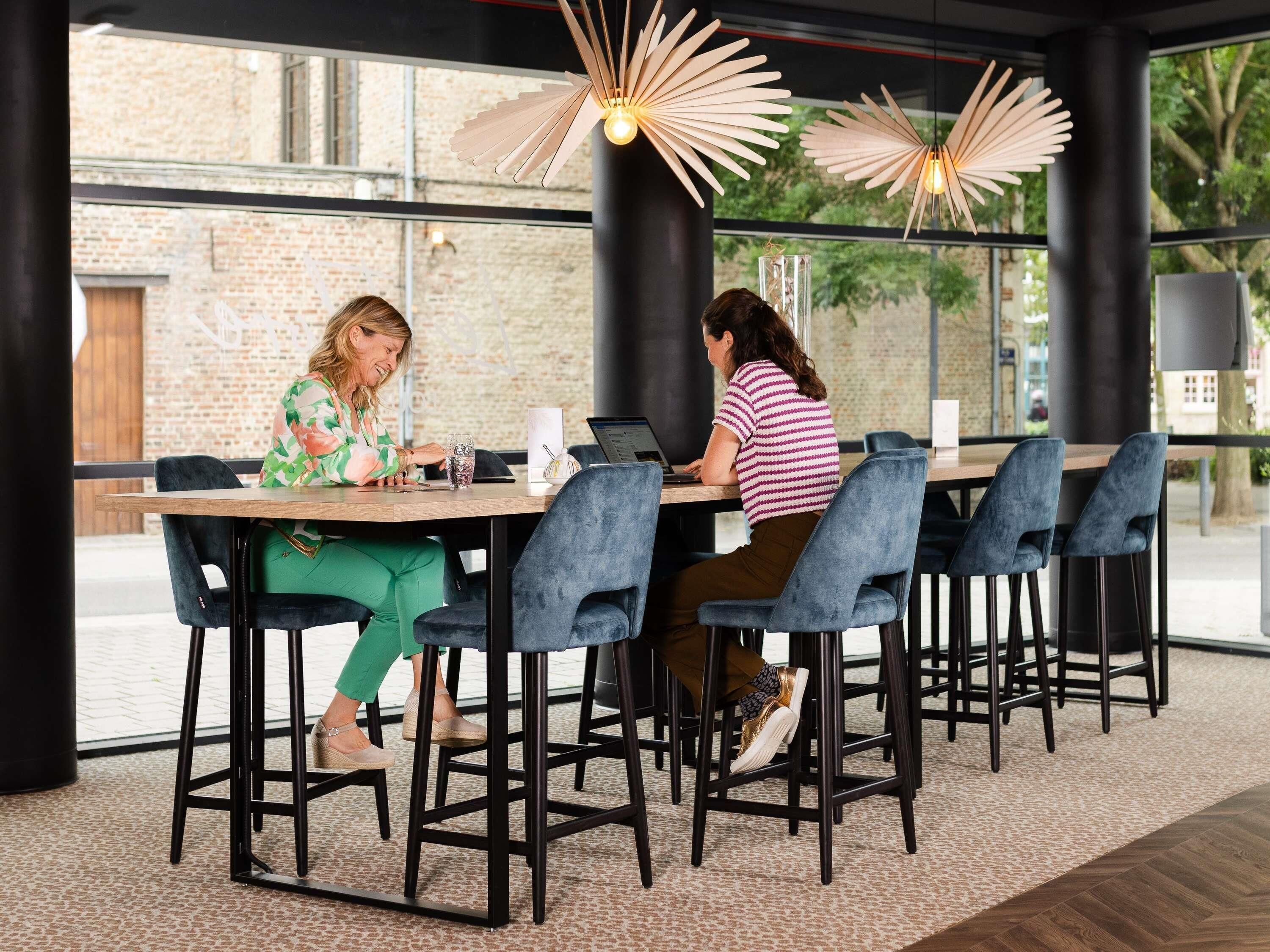 Duas pessoas sentadas à mesa com um laptop, num espaço com amplas janelas e iluminação pendente moderna.