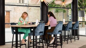 Duas pessoas sentadas à mesa com um laptop, num espaço com amplas janelas e iluminação pendente moderna.
