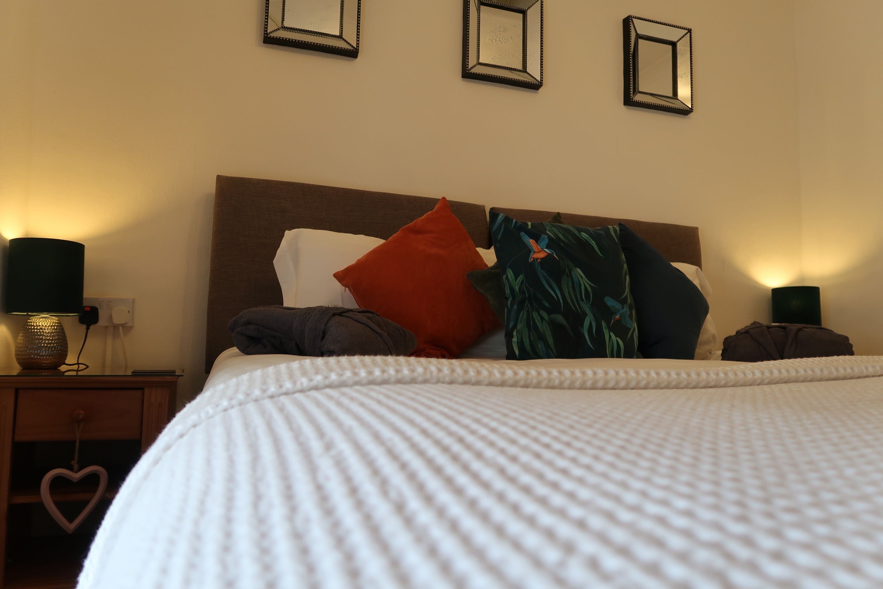 Cama bem arrumada com edredom branco e várias almofadas, mesa de cabeceira com abajur, e parede com três quadros emoldurados.