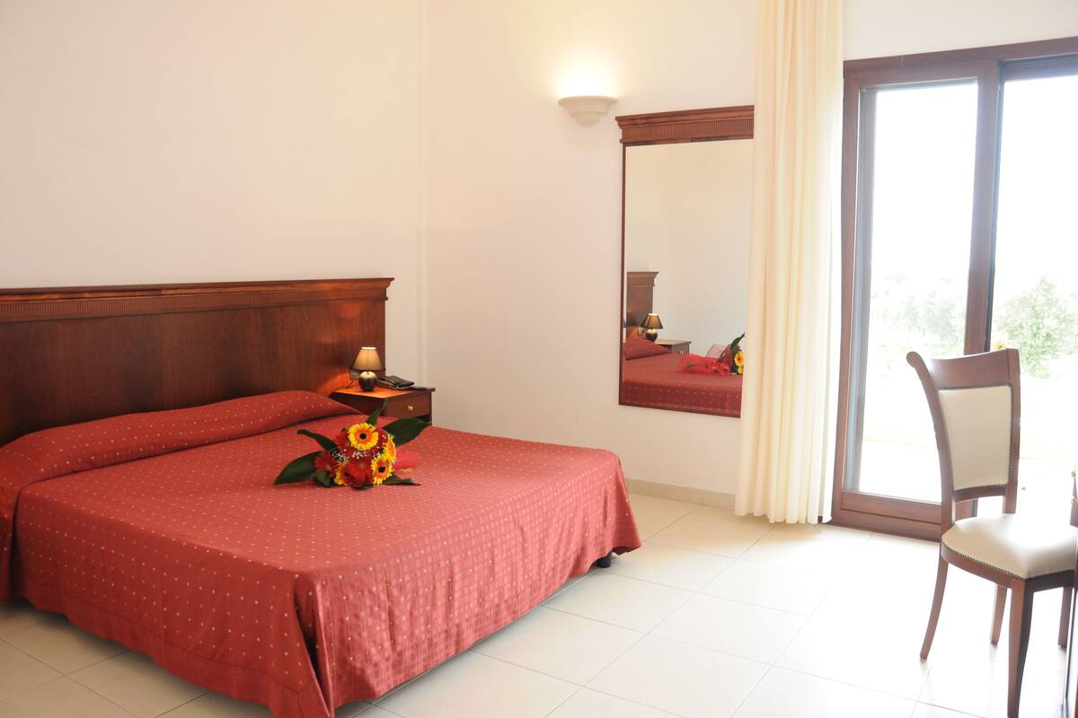 Quarto de hotel com uma cama grande, mesa de cabeceira com abajur, uma cadeira, um espelho e vista para o exterior através de uma porta deslizante.