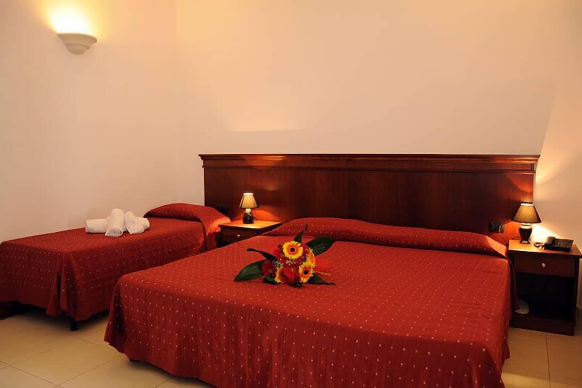Quarto de hotel com duas camas de solteiro e uma cama maior, cada uma com roupa de cama vermelha e cabeceira de madeira. Há luminárias de cabeceira e uma pequena mesa com um abajur.