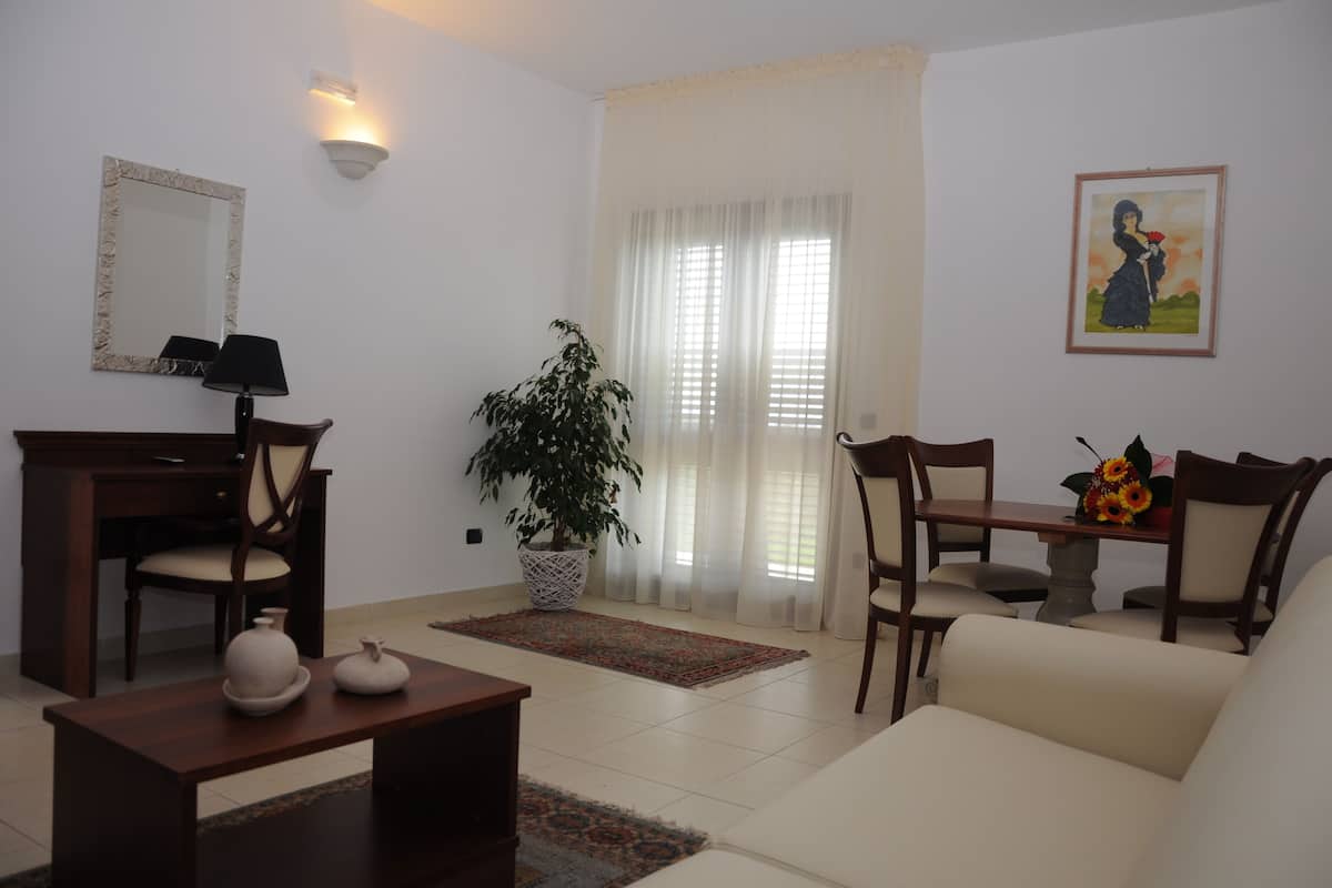 Uma sala de estar com sofá, mesa de centro, mesa de jantar com cadeiras, planta em vaso e um quadro na parede.