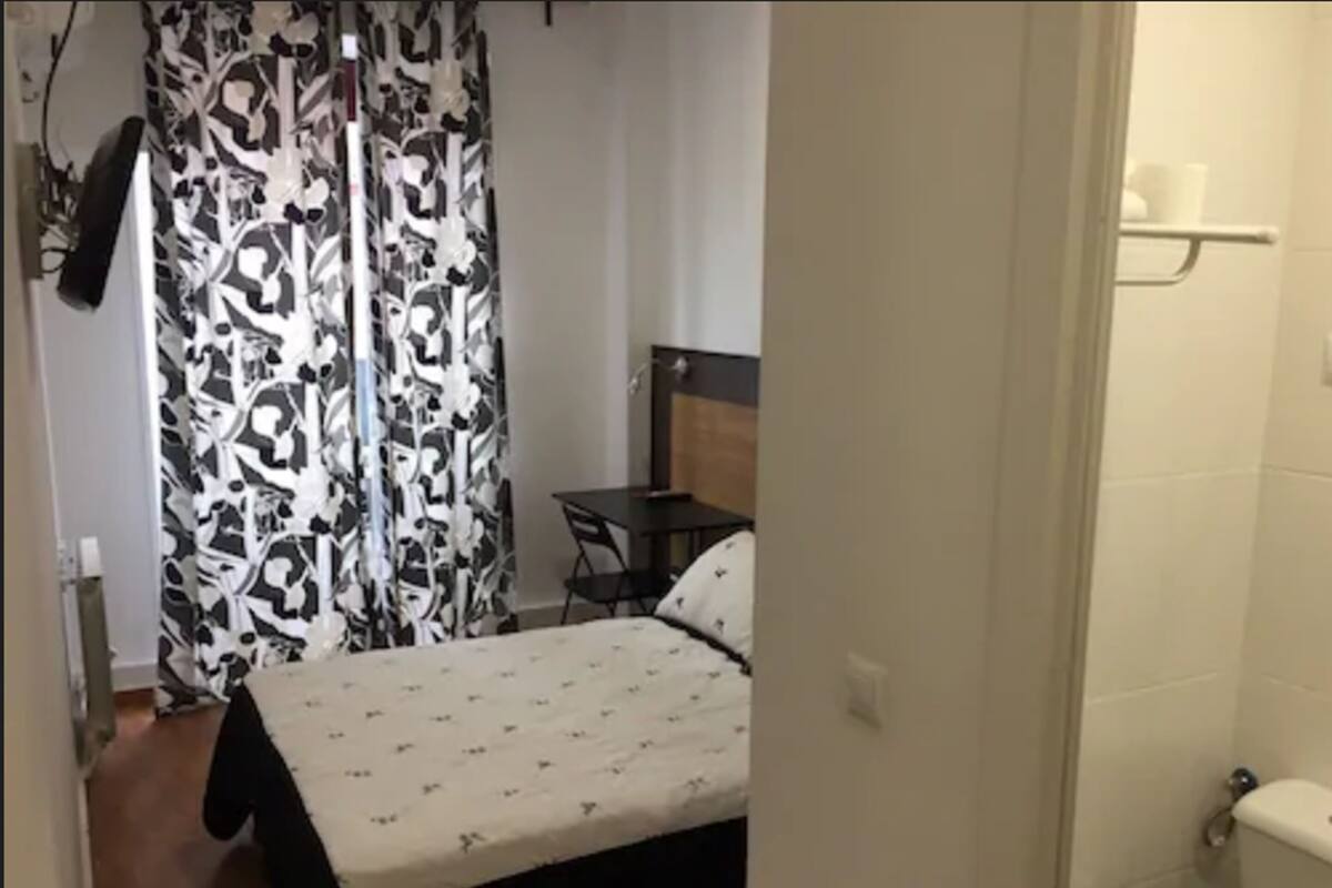 Um quarto com cama, escrivaninha e cadeira. Há uma cortina com padrão em preto e branco.