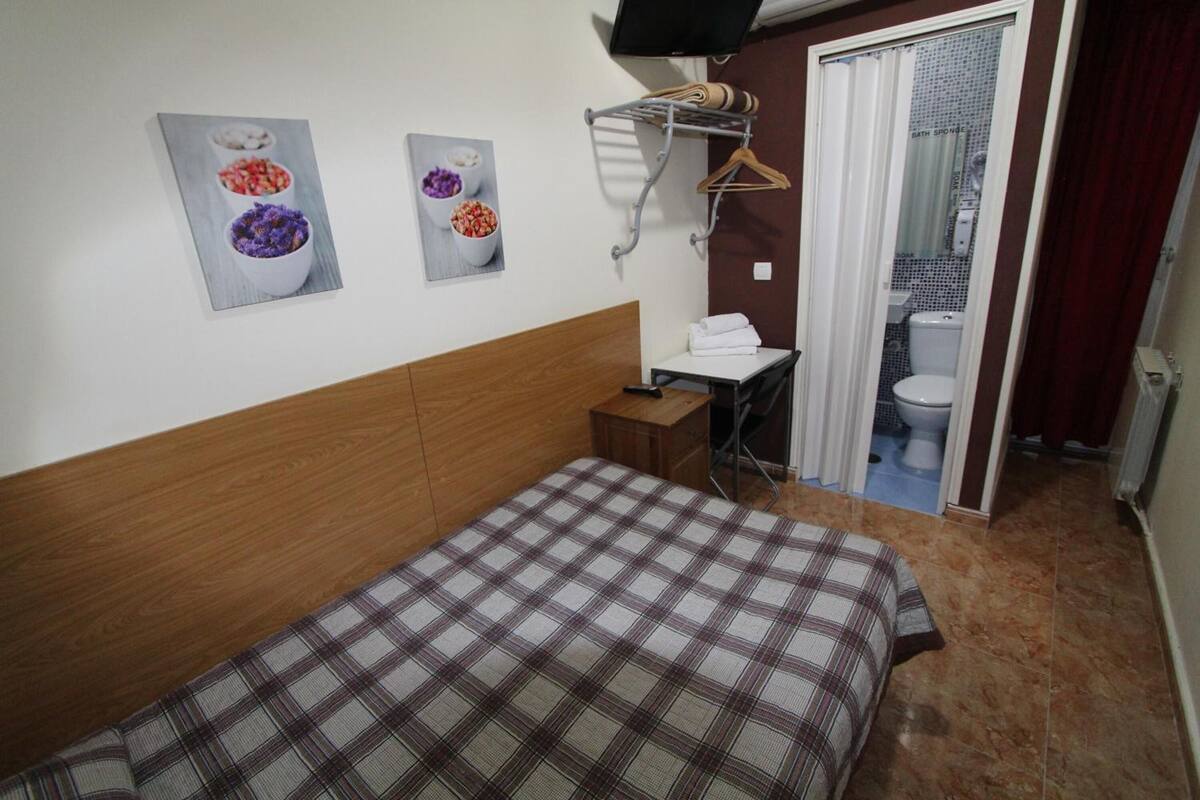Quarto com cama, escrivaninha, cadeira, televisão e banheiro visível por uma porta aberta.