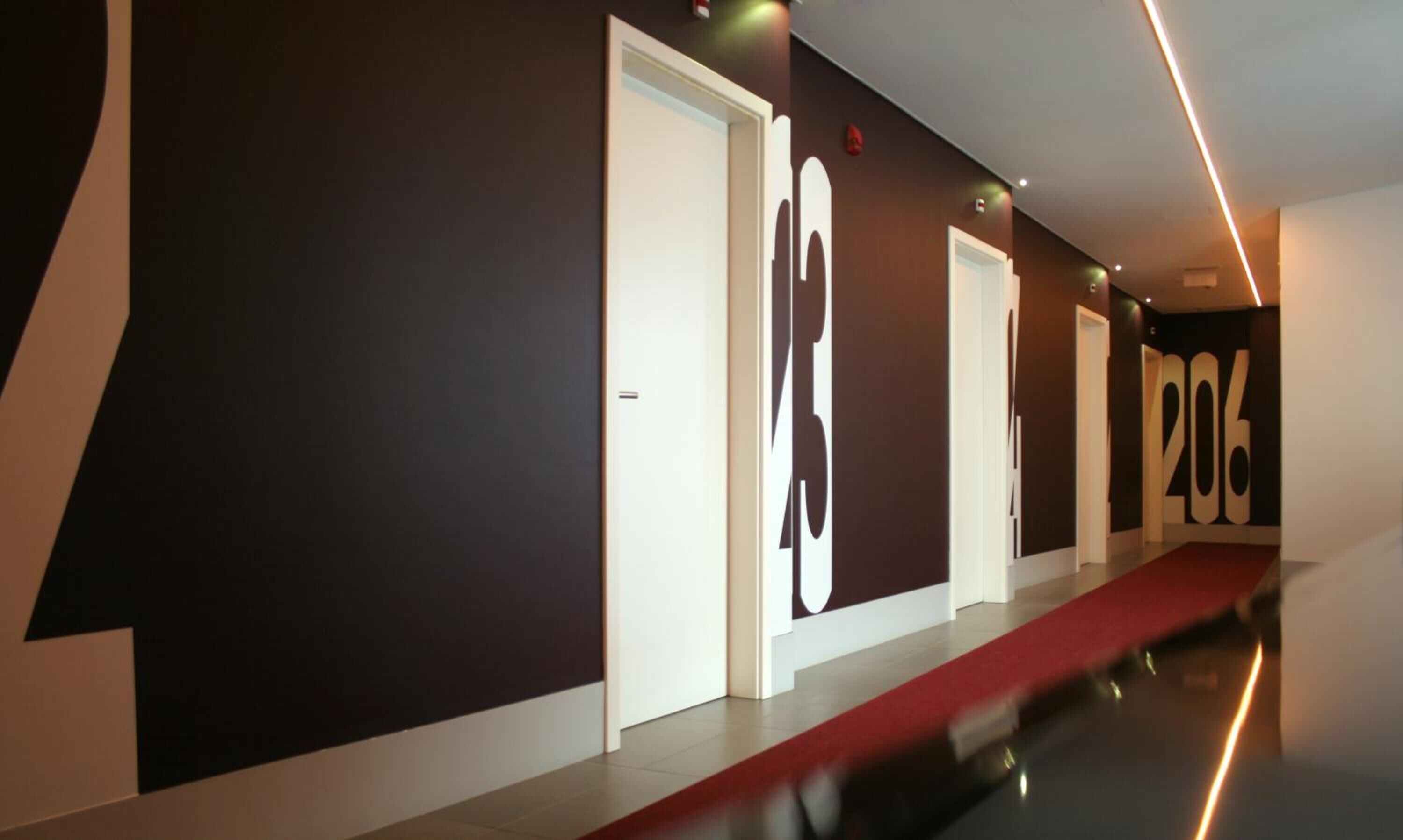 Um corredor moderno com paredes escuras, portas brancas e números grandes indicando os números dos quartos.