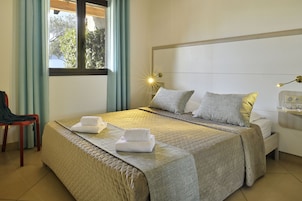Uma cama bem arrumada com uma toalha dobrada, uma mesa de cabeceira com copos e uma janela com cortinas.
