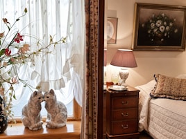 Quarto com cama, mesa de cabeceira com abajur, cômoda e janela com cortinas translúcidas.