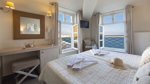 Quarto de hotel com cama, escrivaninha, cadeira, TV e uma grande janela com vista para o mar.