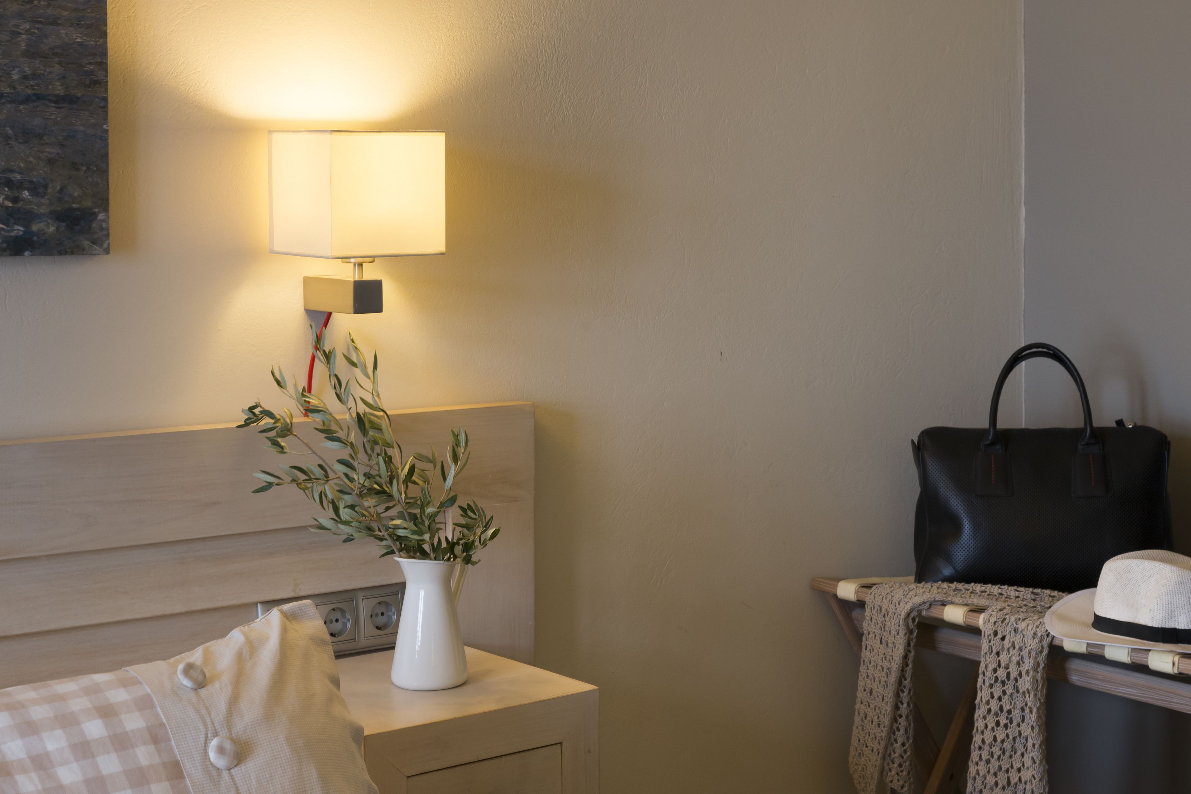 Quarto de hotel bem arrumado, com uma luminária de cabeceira, um vaso branco com uma planta, uma bolsa de mão preta e um chapéu sobre uma mesa de cabeceira de madeira.