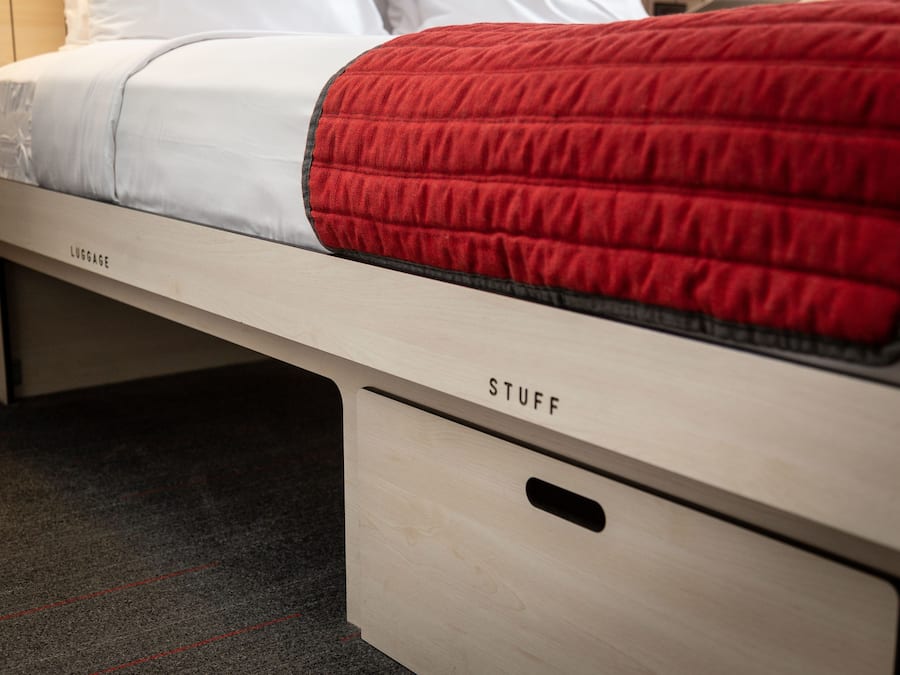 Uma cama com colchão branco e cobertor vermelho, uma cabeceira de madeira com a inscrição 'LUGGAGE' e uma gaveta com a inscrição 'STUFF'.