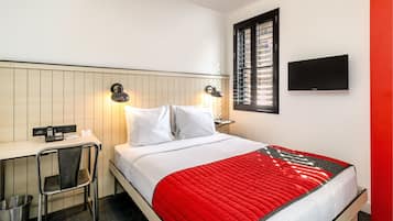 Quarto de hotel com cama, escrivaninha, cadeira, televisão e janela com persianas.