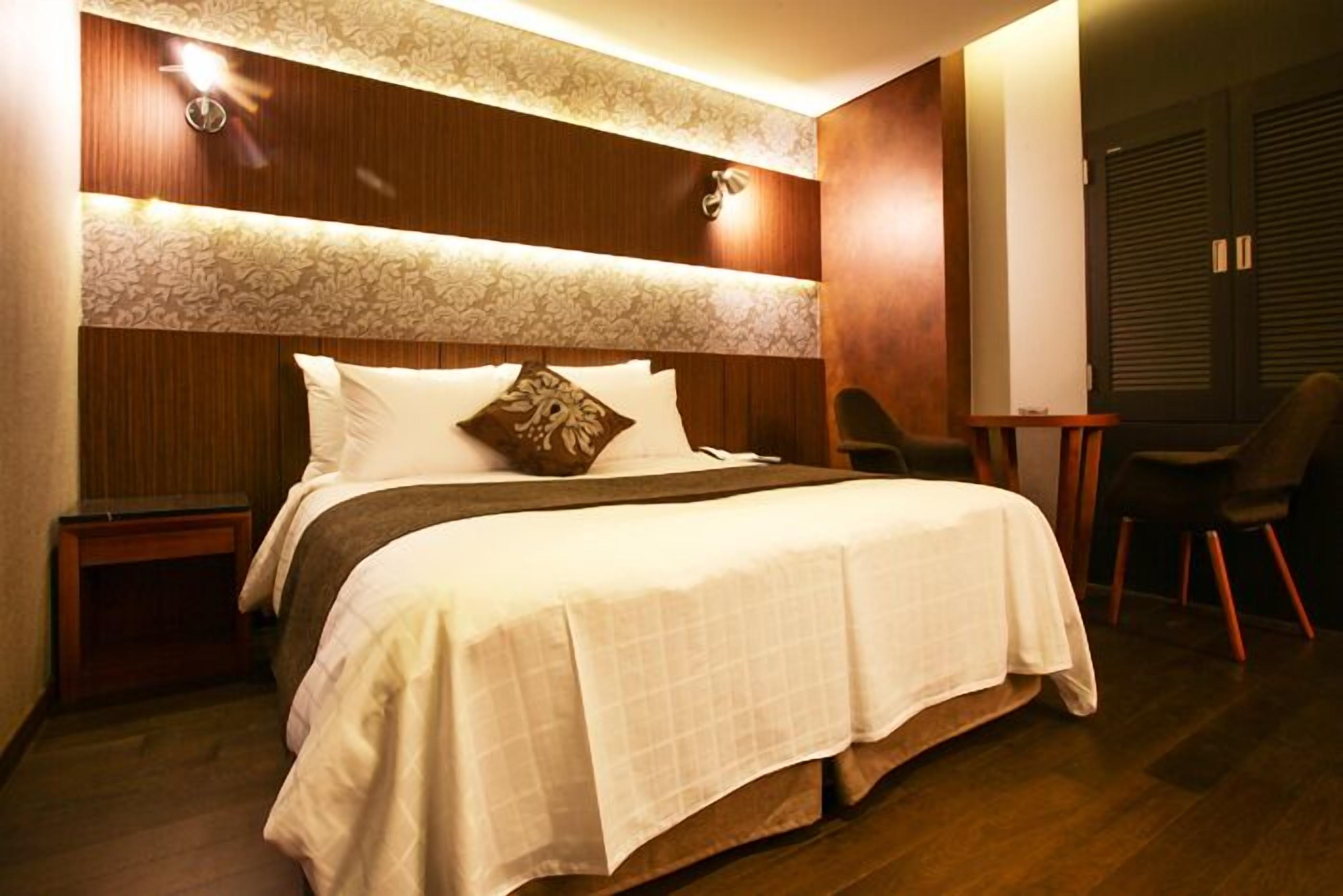 Cama bem arrumada com colcha branca e castanha, um travesseiro decorativo e uma mesinha de cabeceira com abajur. Uma pequena mesa de madeira e duas cadeiras estão posicionadas à direita da cama.