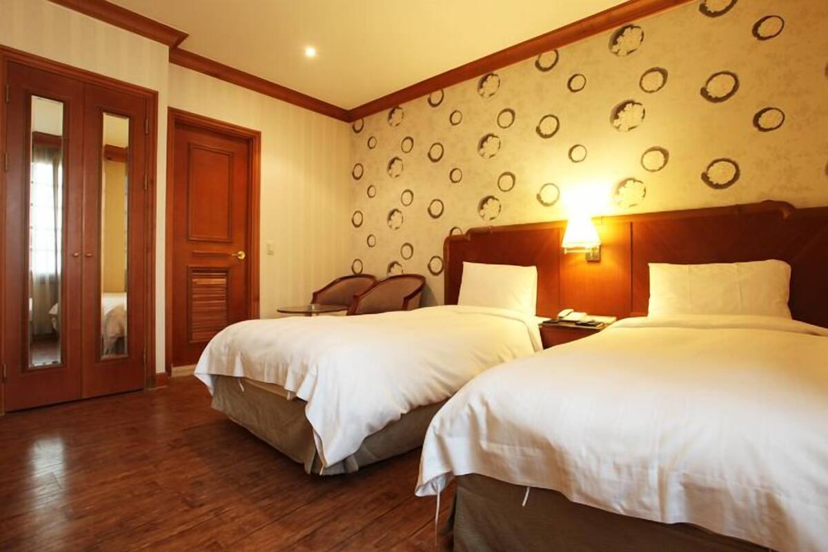 Quarto de hotel com duas camas, soalho de madeira, parede estampada e porta com espelho.