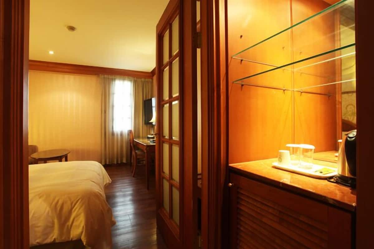 Quarto de hotel com uma cama, uma escrivaninha, uma cadeira, um aparador de vidro e uma mesinha com chaleira e chávenas.