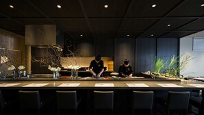 Uma cozinha moderna com chefs preparando comida, um balcão comprido com papéis e decoração minimalista, incluindo galhos e flores brancas.