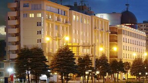 Um hotel com vários andares e varandas, um edifício em forma de cúpula à direita e luminárias de rua à frente.