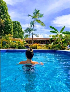 Uma pessoa nada em uma piscina com vista para um edifício rodeado por vegetação tropical.