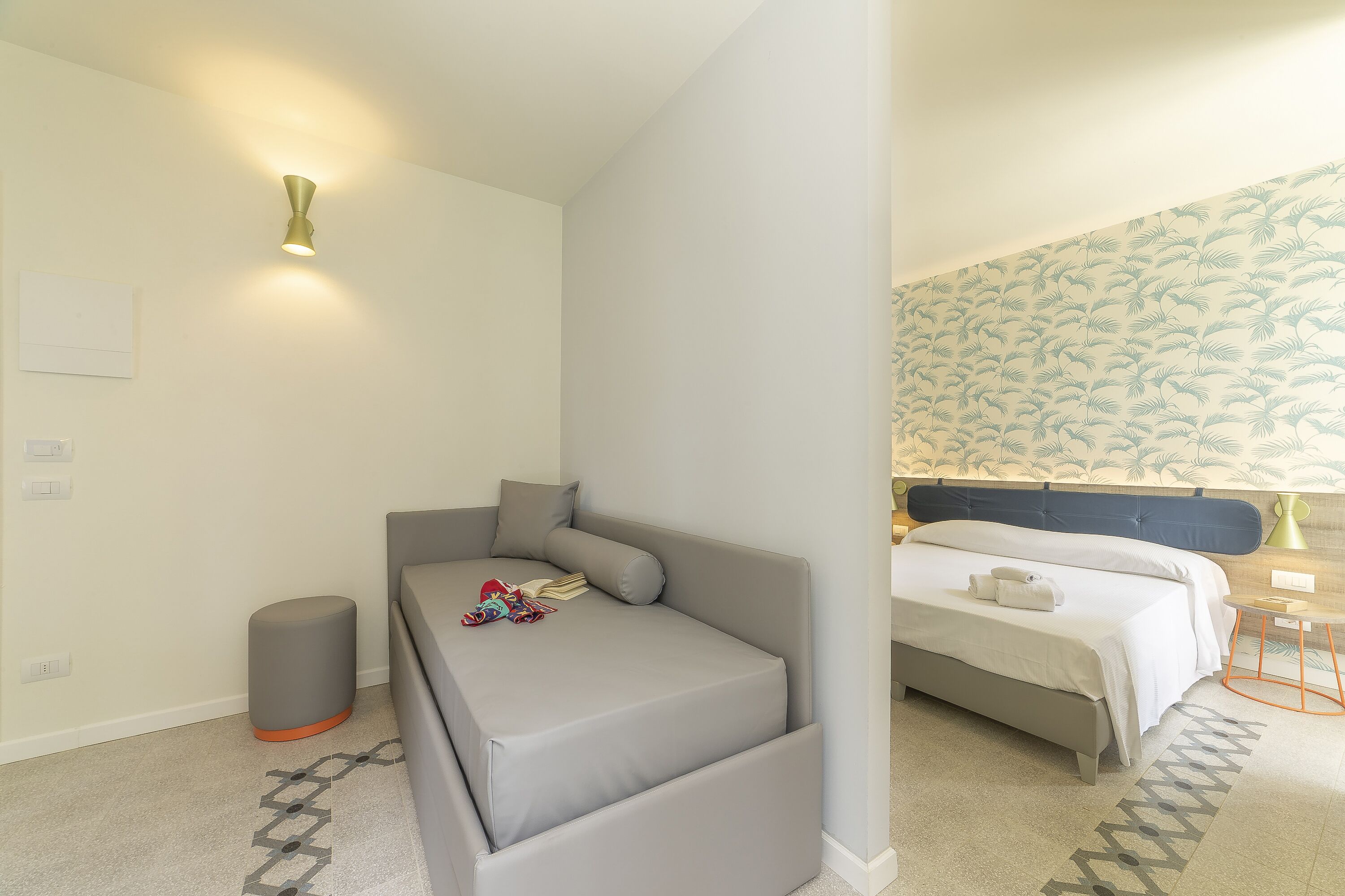 Quarto moderno com cama-sofá, cama de casal e papel de parede estampado, além de uma mesinha de cabeceira.