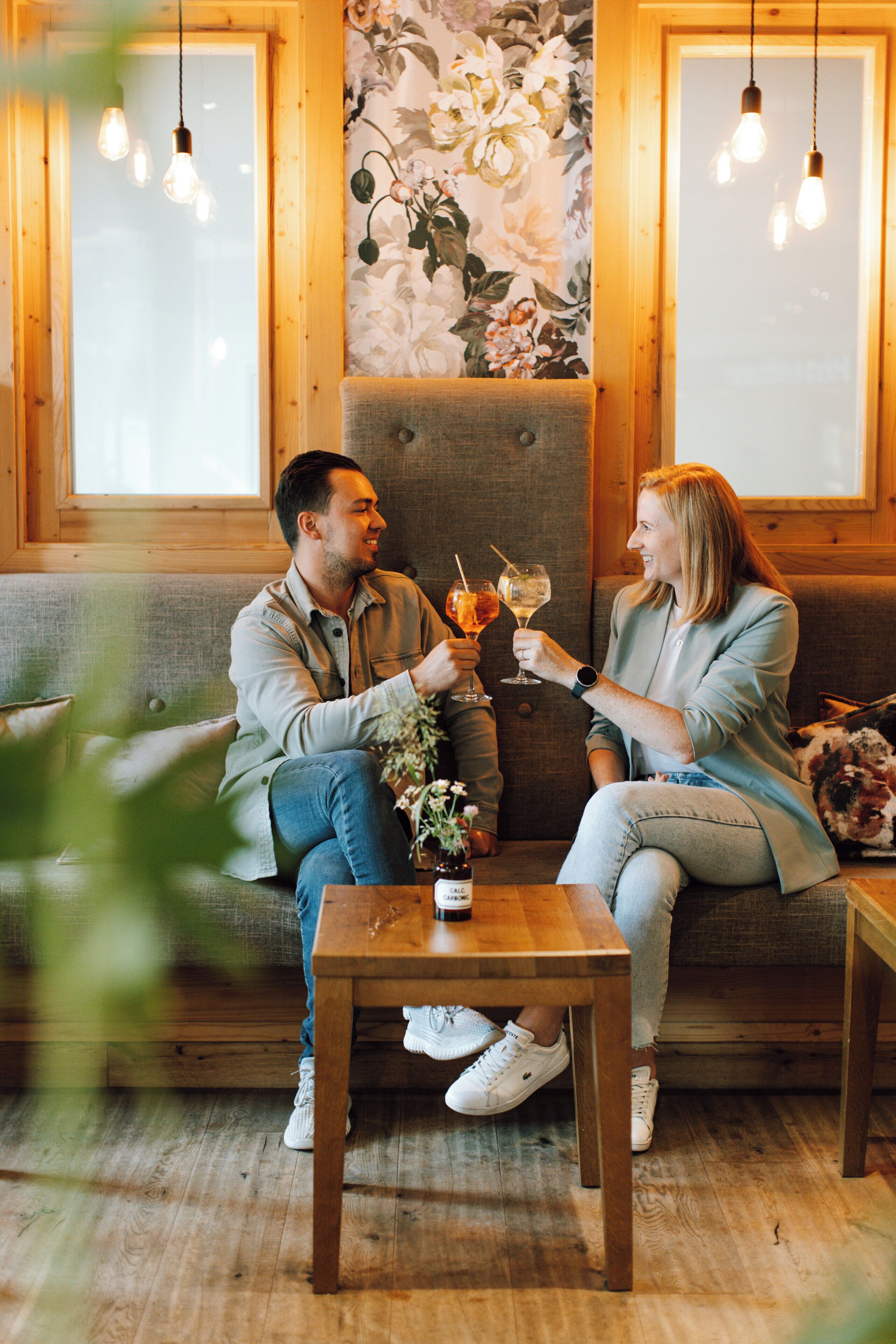 Interior de madeira com sofá e uma pequena mesa, duas pessoas sentadas brindando com bebidas.