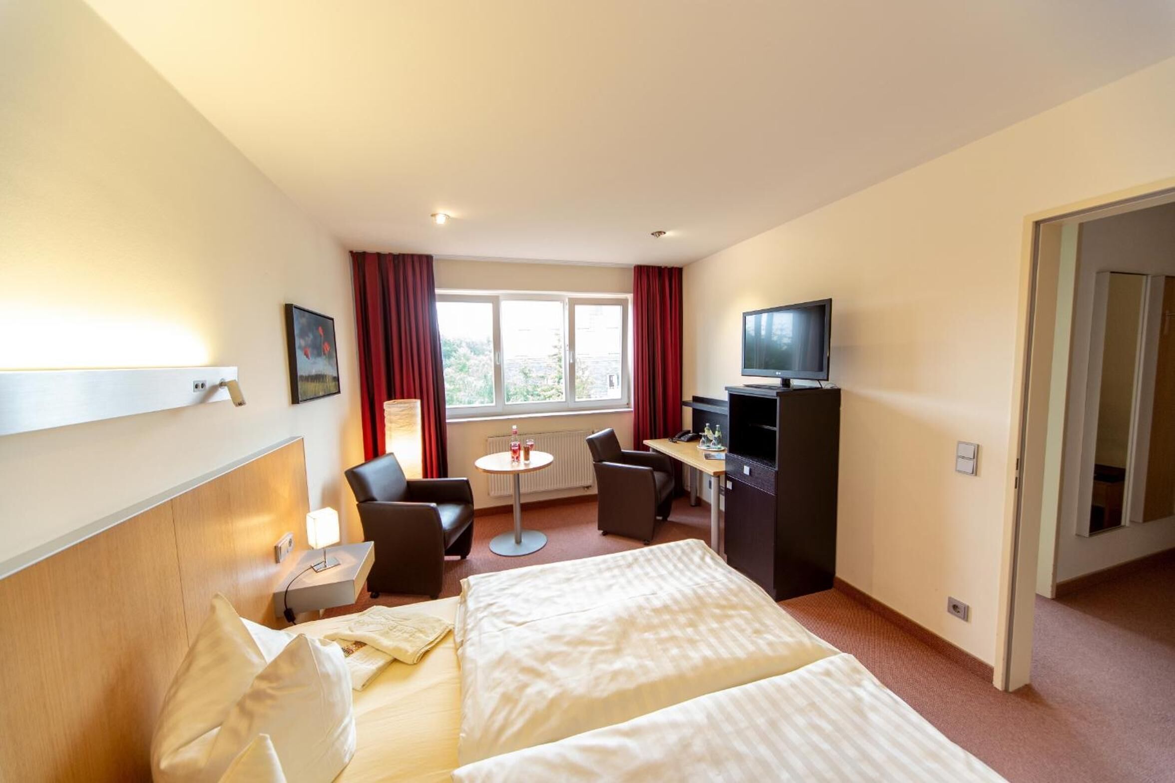 Quarto de hotel com cama, escrivaninha, cadeira, televisão e janela com cortinas.