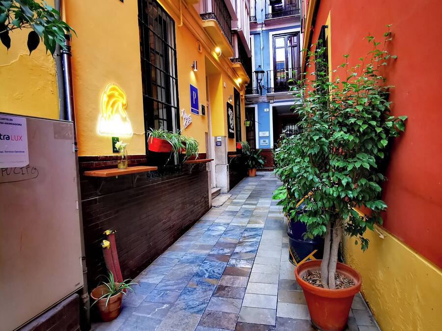 Uma viela estreita com um prédio amarelo, um letreiro de neon e plantas em vasos.