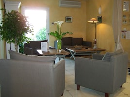 Um saguão de hotel com poltronas cinzas, um sofá, uma mesa de centro, um vaso com flores, um vaso de planta e um abajur.