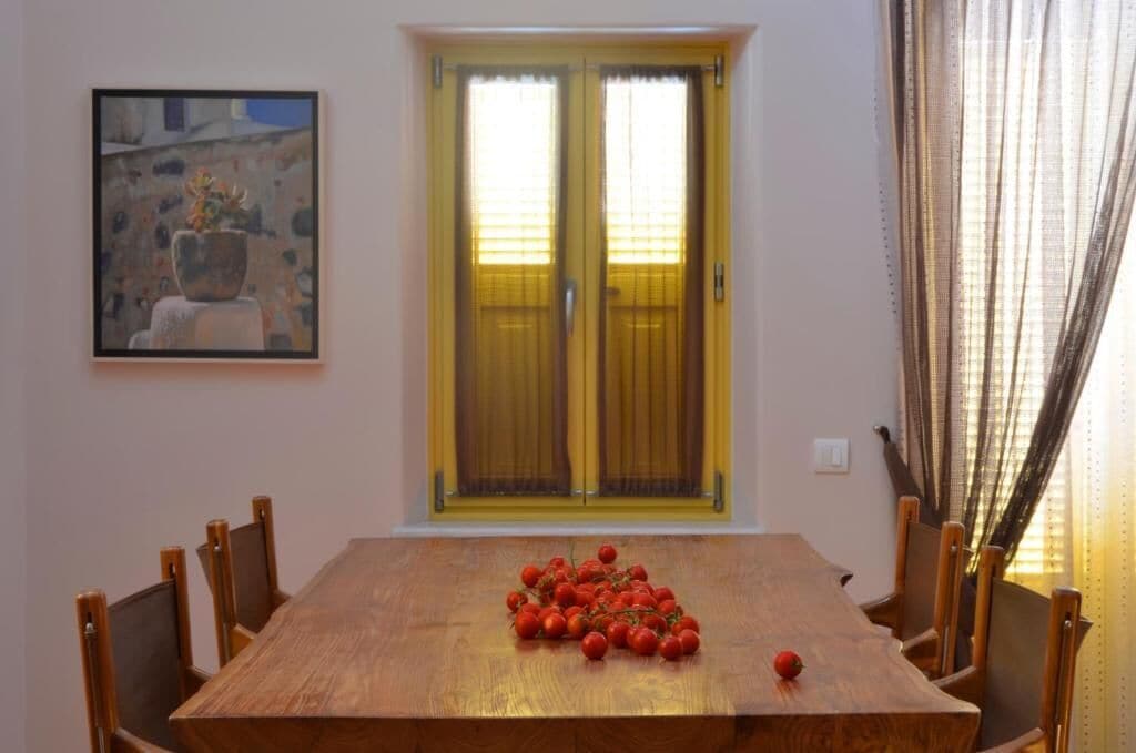 Uma sala de jantar com uma mesa de madeira, cadeiras, um quadro na parede e uma janela com cortinas.