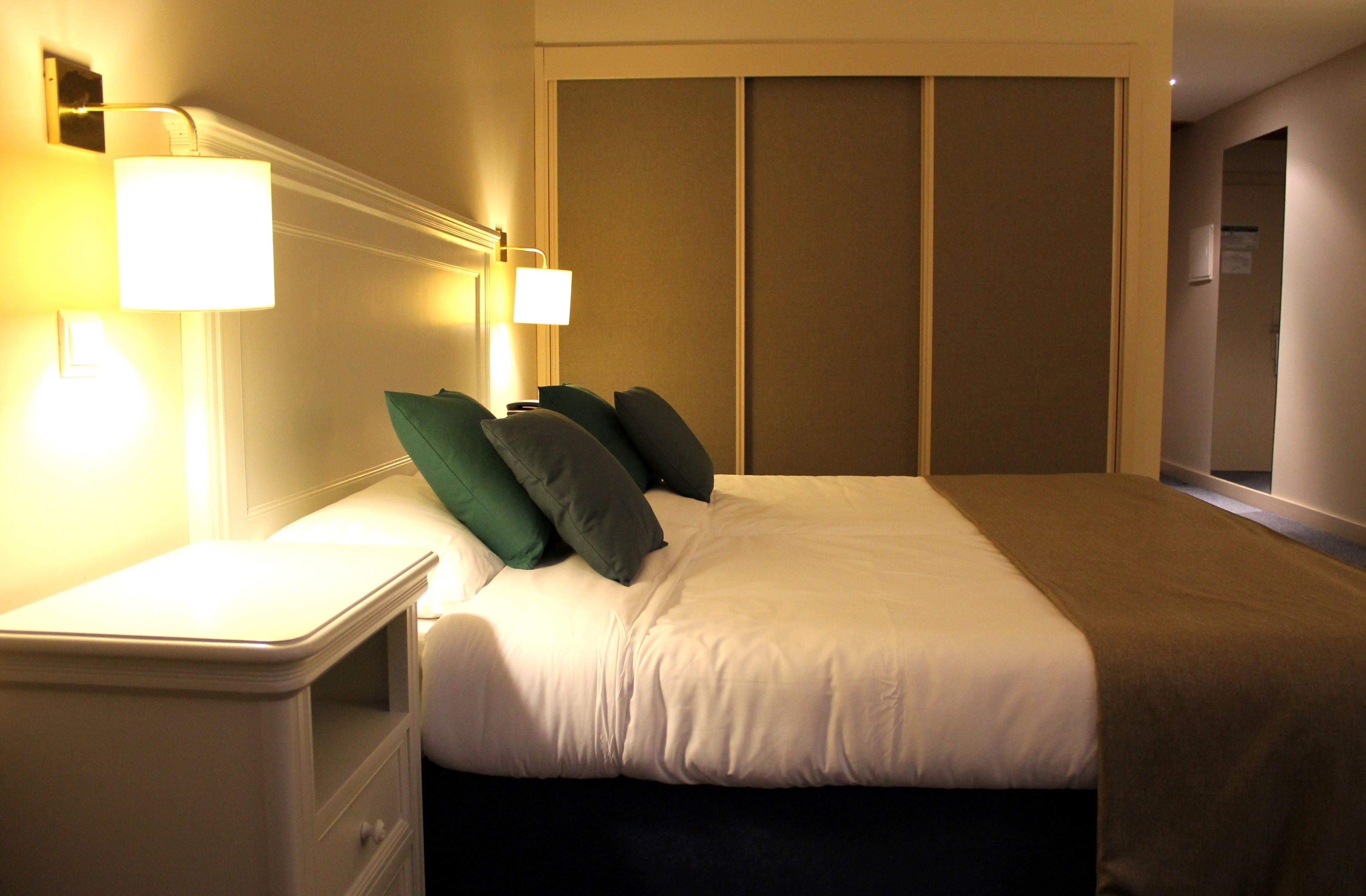 Cama bem arrumada com roupa de cama branca e almofadas verdes, mesa de cabeceira com abajur e um guarda-roupa com portas de correr ao fundo.