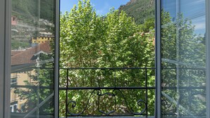 Visão pela janela com grade de metal, mostrando uma árvore verde e um morro com edifícios ao fundo.