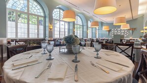 Uma sala de jantar com mesas redondas preparadas para refeição, amplas janelas em arco e luminárias pendentes.