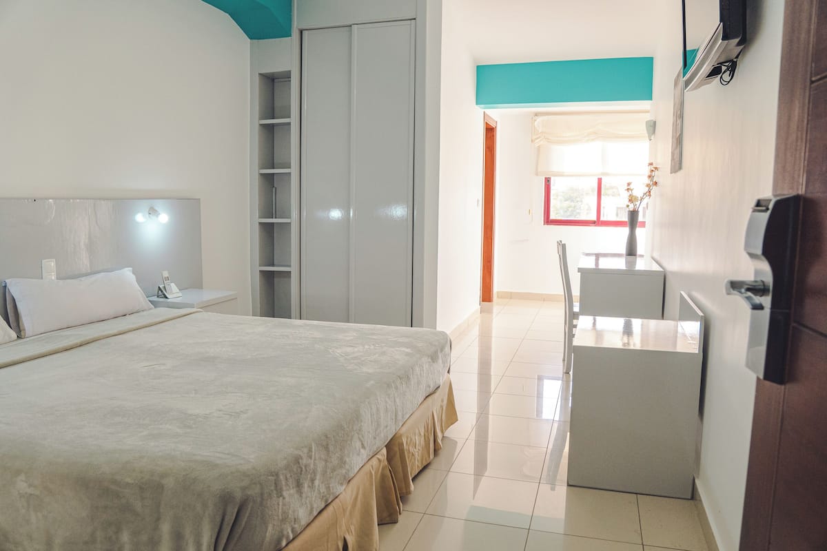 Quarto de hotel com cama, escrivaninha, guarda-roupa e janela com cortinas.