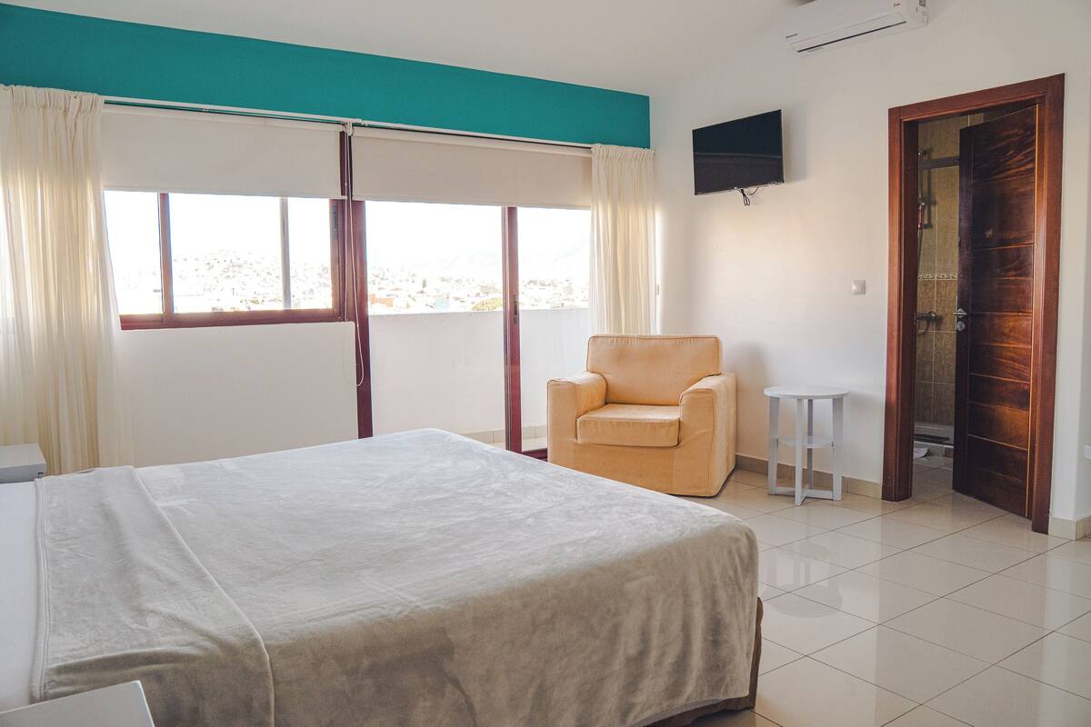 Quarto de hotel com cama, uma cadeira, uma mesinha, TV fixada na parede e porta de acesso a um banheiro.
