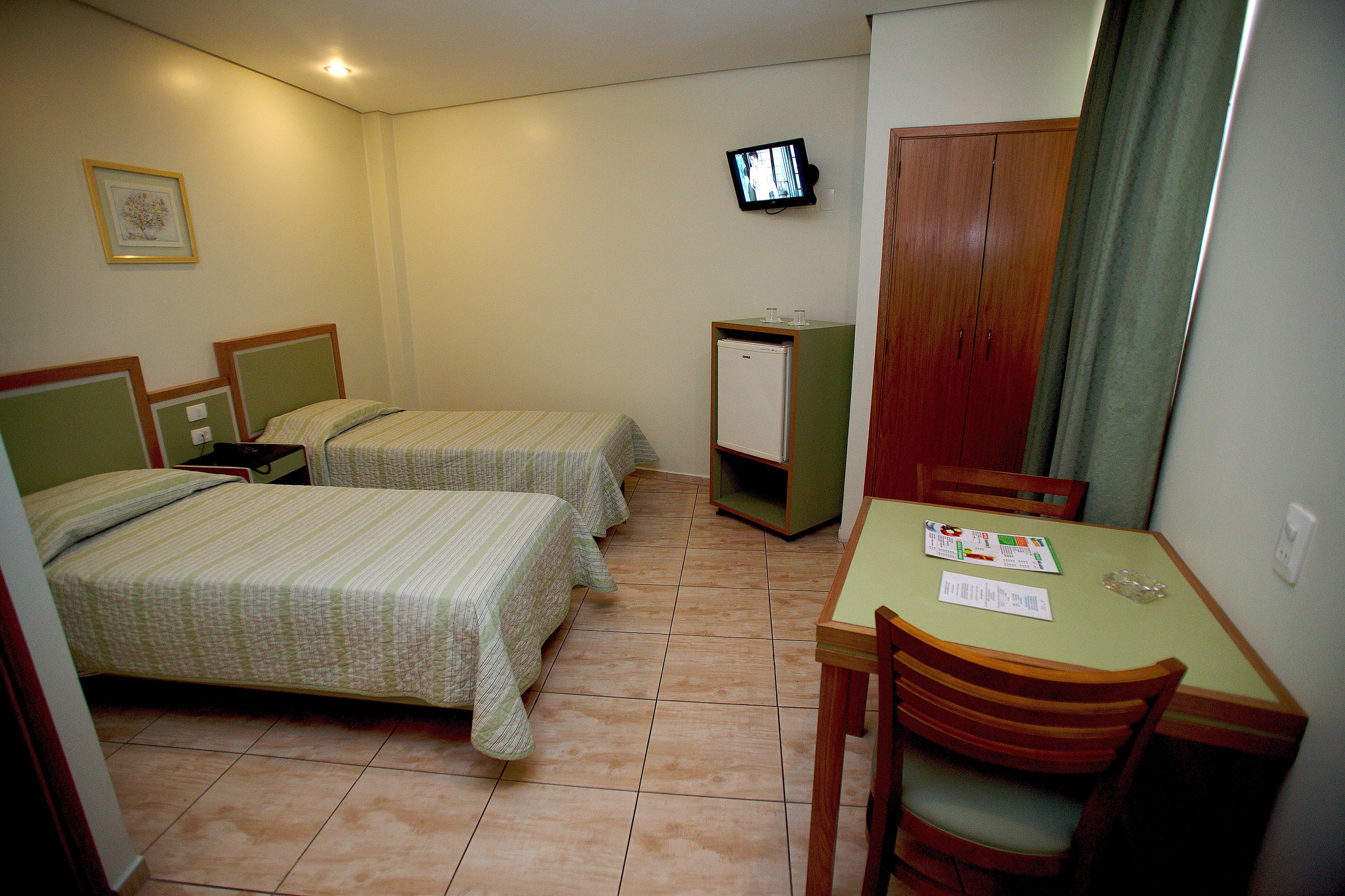 Quarto de hotel pequeno com duas camas de solteiro, uma escrivaninha, televisão e guarda-roupa.