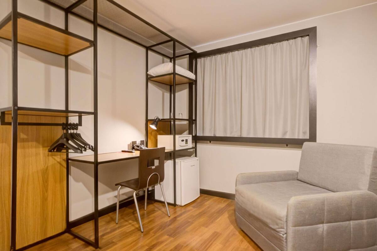 Quarto pequeno com escrivaninha de madeira, uma cadeira, uma cama e janela com cortinas.