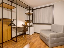 Quarto pequeno com escrivaninha de madeira, uma cadeira, uma cama e janela com cortinas.