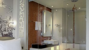 Quarto de hotel moderno com uma cama, banheiro com box de vidro, toucador branco com bancada preta e um espelho grande.