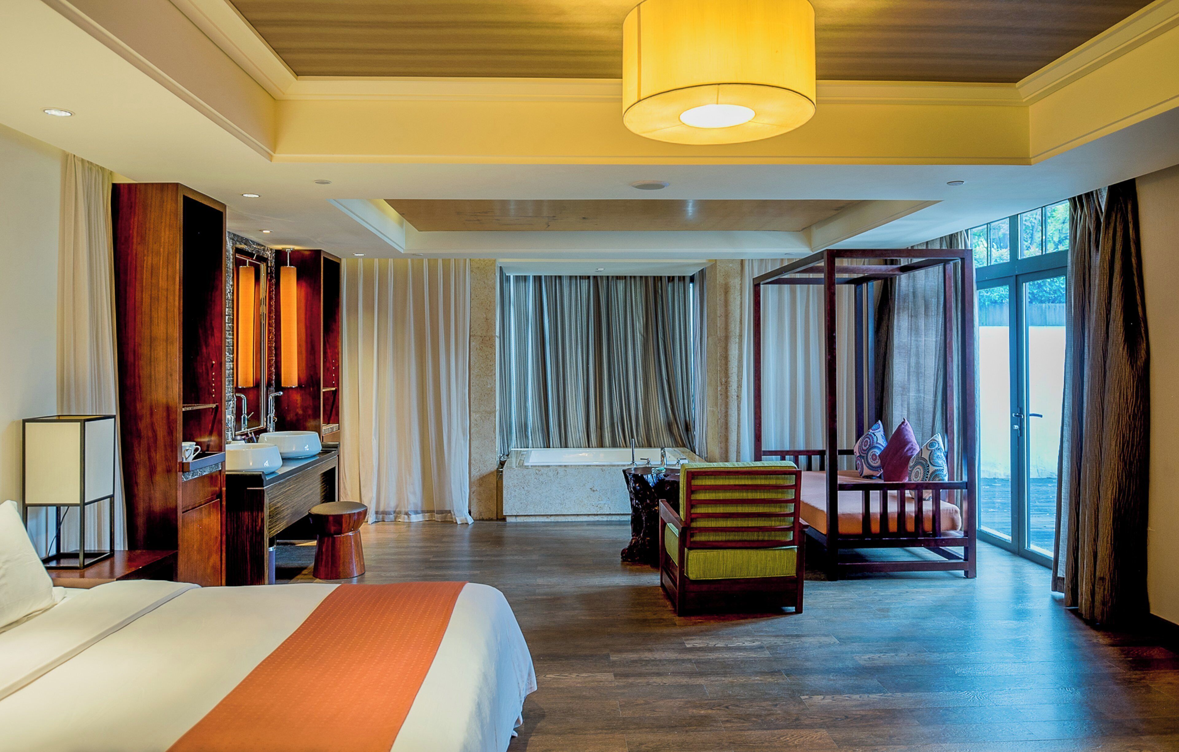 Quarto de hotel com uma cama grande, área de estar com uma cadeira, escrivaninha com abajur, janela com cortinas e luminária no teto.
